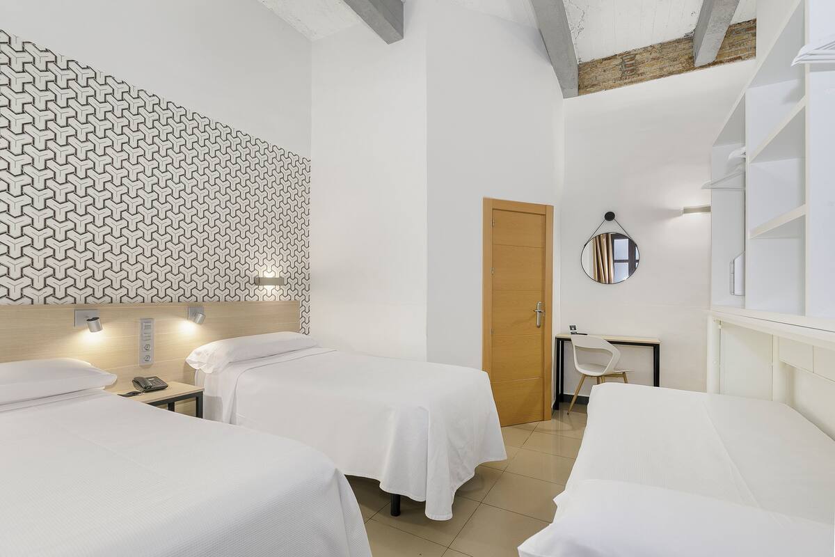 Quarto de hotel com duas camas, uma escrivaninha, uma cadeira, uma porta de madeira e parede estampada.
