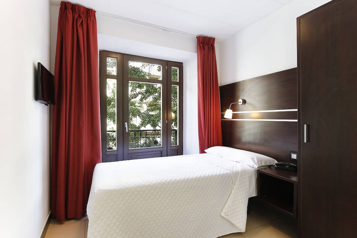 Um quarto com cama, mesa de cabeceira, televisão na parede e janela com cortinas.