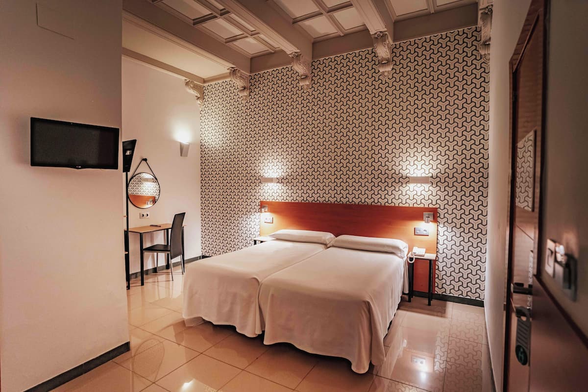 Quarto de hotel com cama de casal, escrivaninha com cadeira, TV fixada na parede e papel de parede estampado.