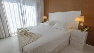 Cama bem arrumada com edredom branco e travesseiros, mesa de cabeceira com abajur e telefone.