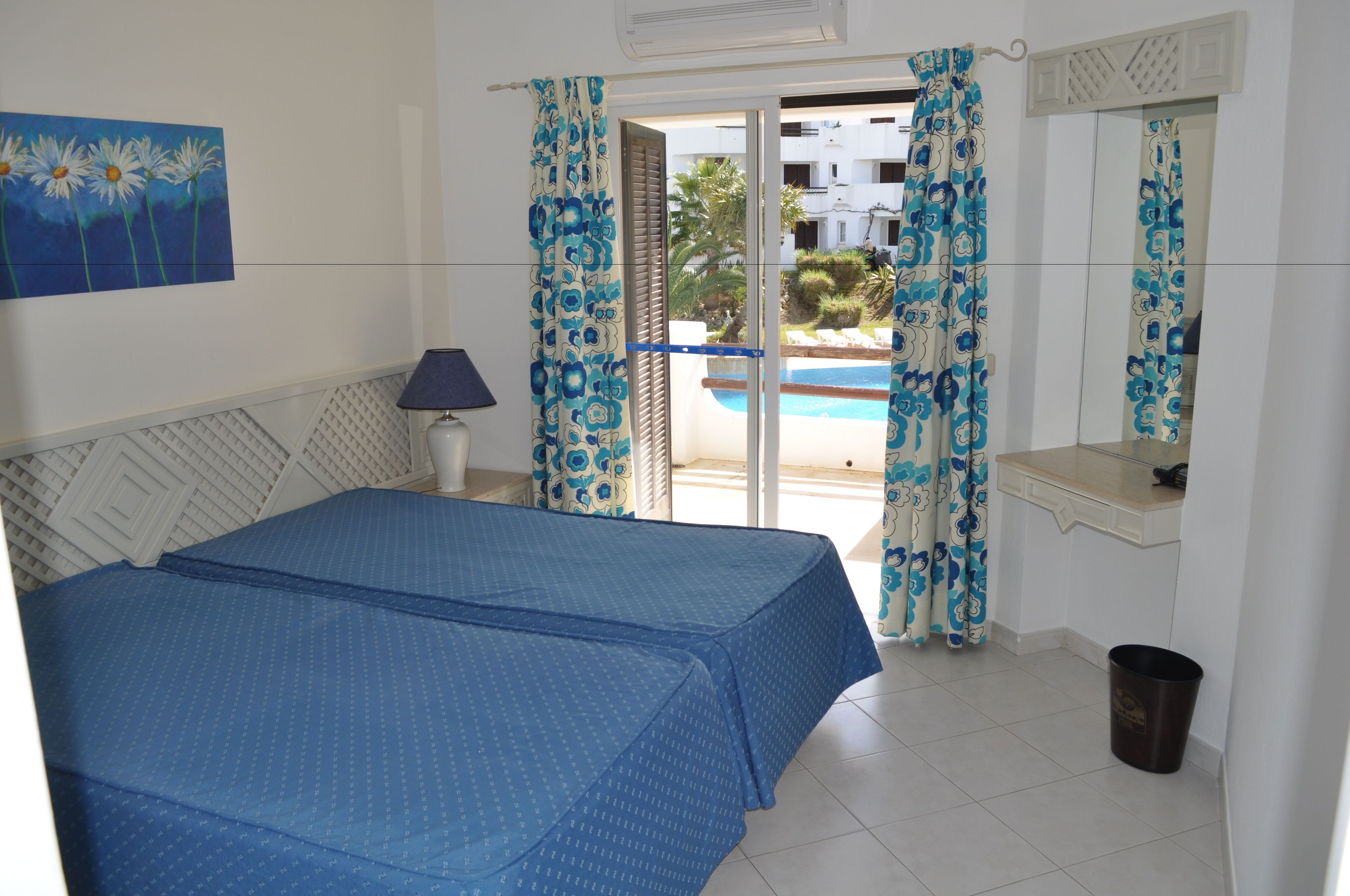 Quarto com cama, mesa de cabeceira, luminária, janela com cortinas, espelho e vista para uma piscina e edifícios.