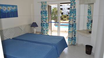 Quarto com cama, mesa de cabeceira, luminária, janela com cortinas, espelho e vista para uma piscina e edifícios.