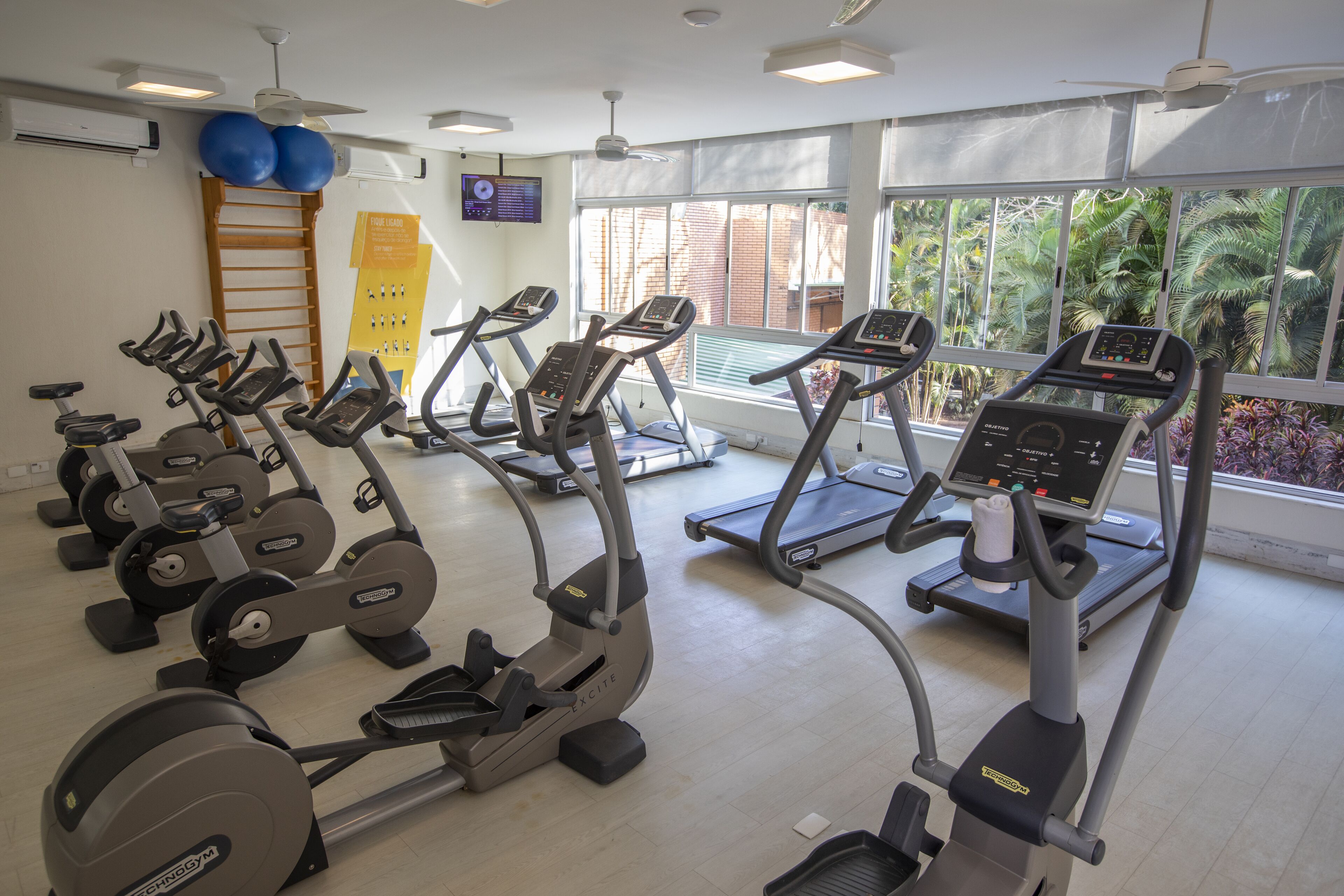 Uma academia com vários equipamentos de cardio, uma tela de televisão e amplas janelas com vista para o verde.