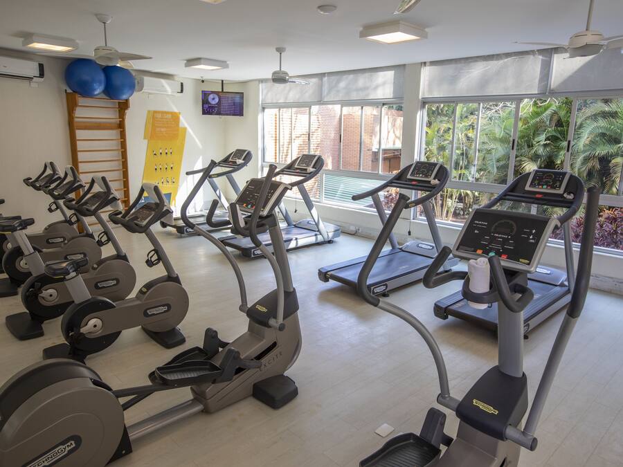 Uma academia com vários equipamentos de cardio, uma tela de televisão e amplas janelas com vista para o verde.