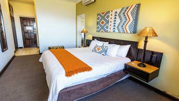 Quarto de hotel com uma cama grande, mesa de cabeceira com abajur, ar condicionado fixado na parede e porta que dá para outro aposento.