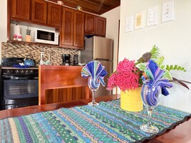 Cozinha com armários de madeira, micro-ondas, fogão e geladeira. Há um jogo americano colorido em tear no balcão, um vaso com flores e um copo com um guardanapo azul e roxo.