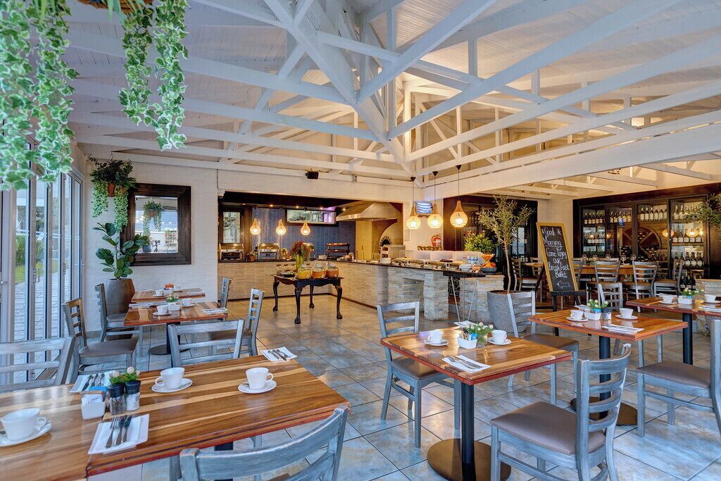 Um café amplo com mesas e cadeiras de madeira, área de bar e plantas penduradas.