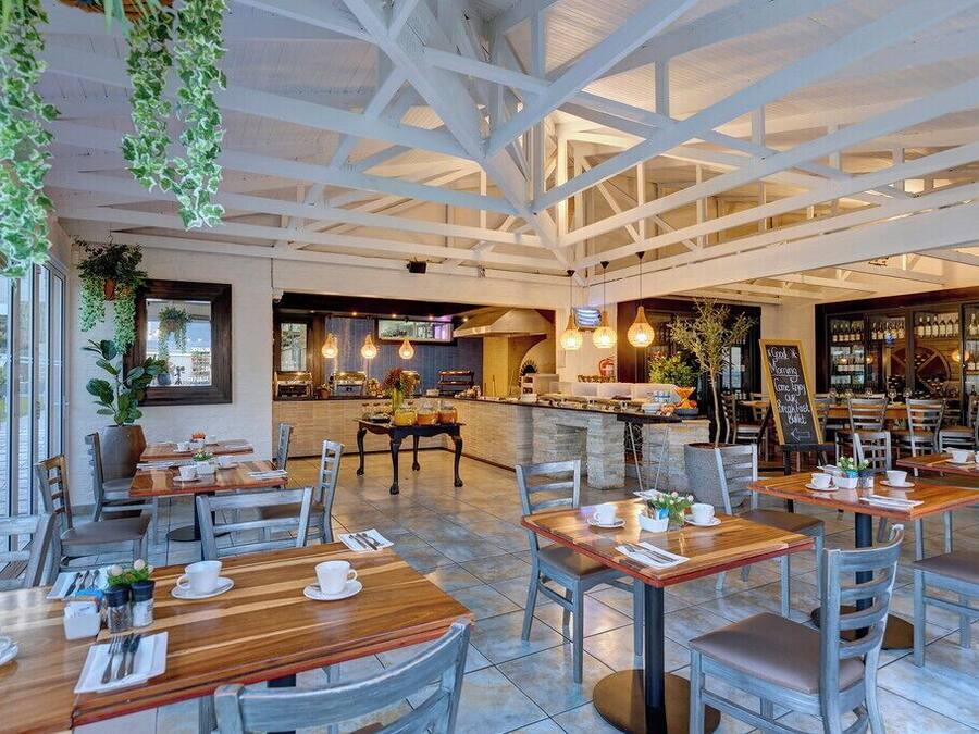 Um café amplo com mesas e cadeiras de madeira, área de bar e plantas penduradas.