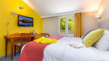 Quarto de hotel com cama, escrivaninha, cadeira, televisão e janela com cortinas.