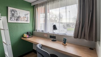 Um quarto com parede verde texturizada, um quadro emoldurado, uma escrivaninha com chaleira, uma luminária e uma janela com cortinas.
