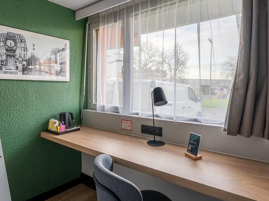 Um quarto com parede verde texturizada, um quadro emoldurado, uma escrivaninha com chaleira, uma luminária e uma janela com cortinas.