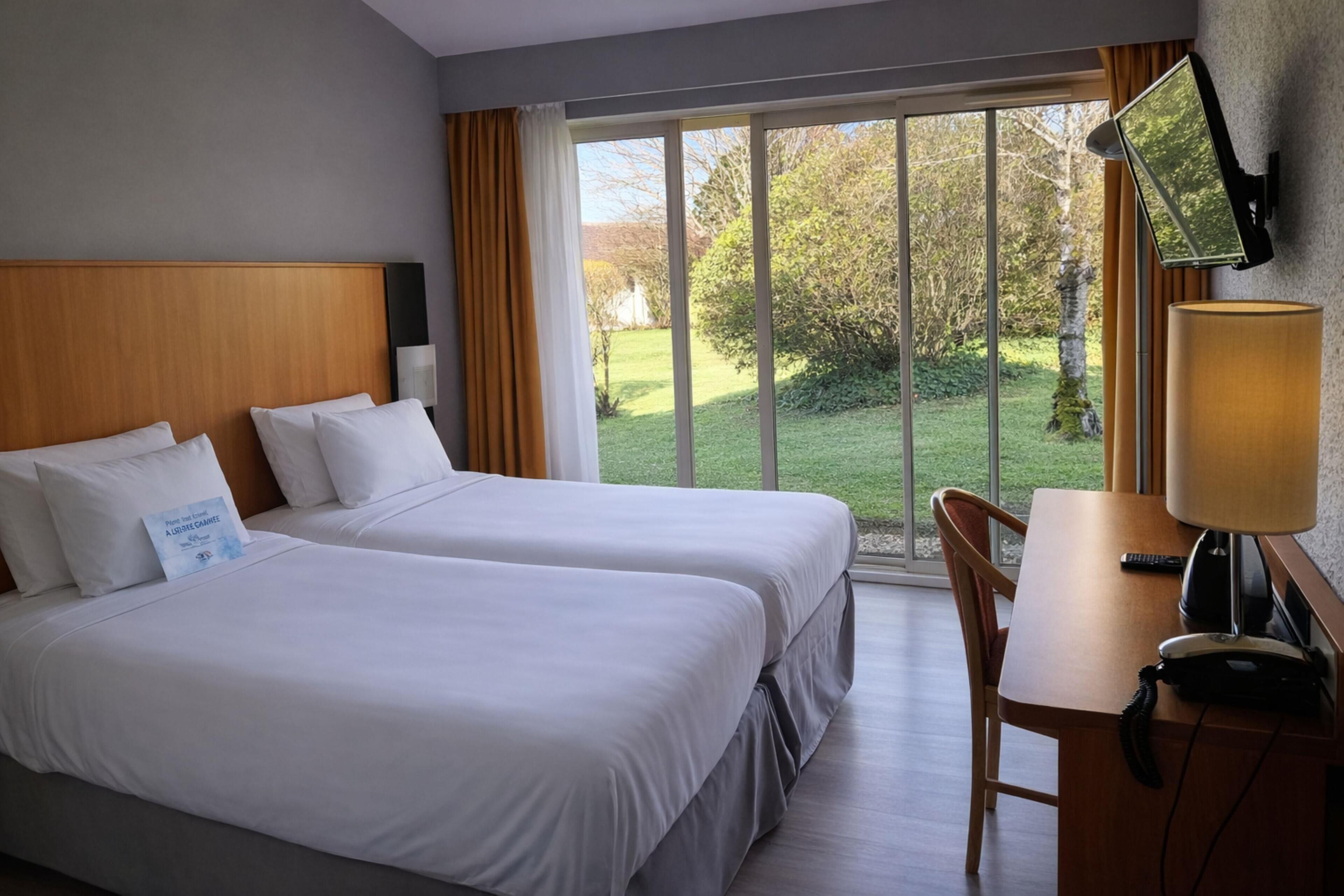 Quarto de hotel com duas camas, uma escrivaninha com abajur, TV fixada na parede e uma grande janela com vista para o verde.
