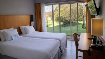 Quarto de hotel com duas camas, uma escrivaninha com abajur, TV fixada na parede e uma grande janela com vista para o verde.