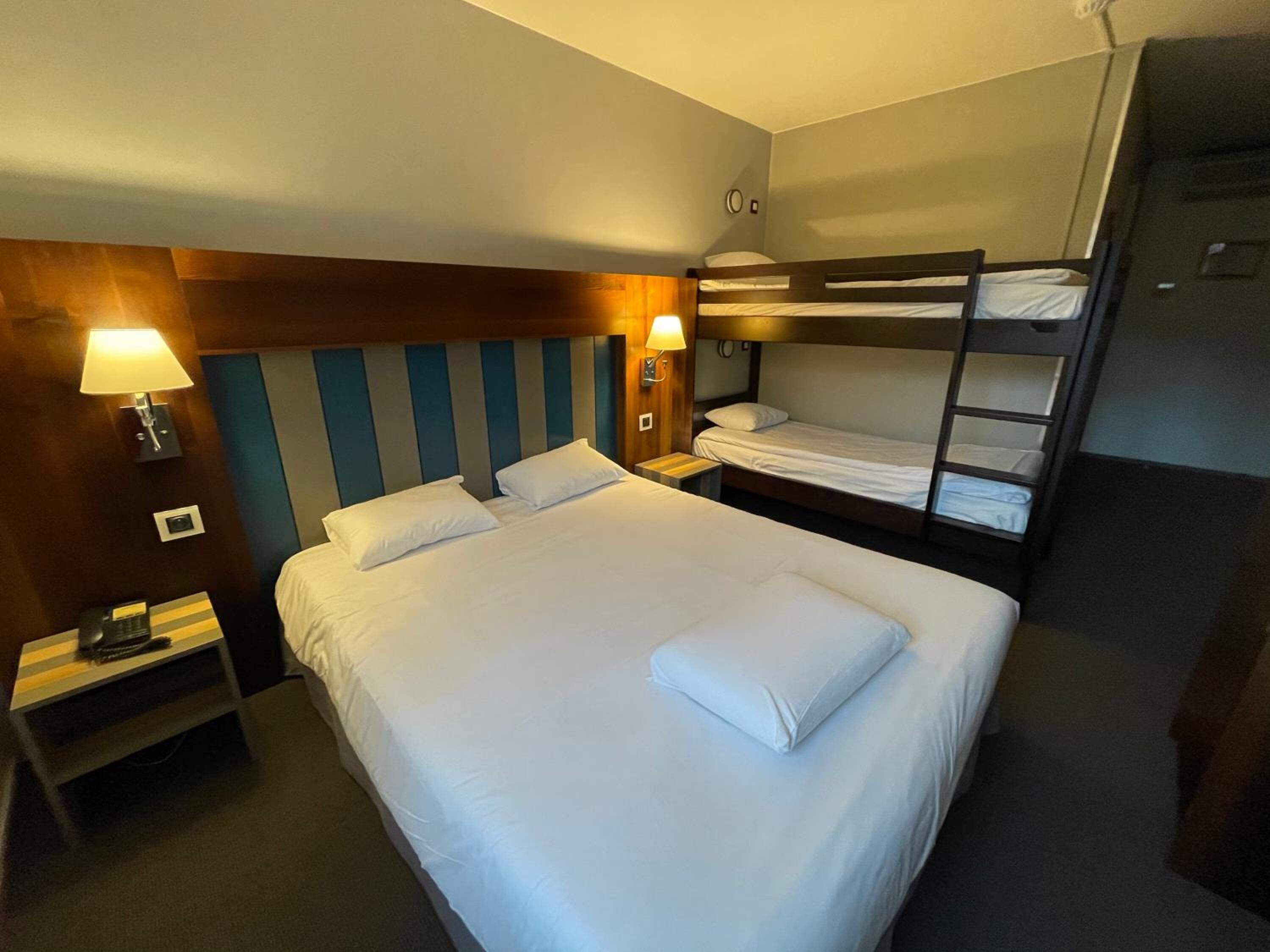 Quarto de hotel com uma cama grande, duas mesas de cabeceira, uma cama beliche e uma mesinha de apoio.