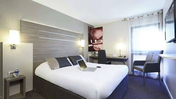 Quarto de hotel com uma cama grande, uma escrivaninha com um laptop, uma cadeira e uma televisão.