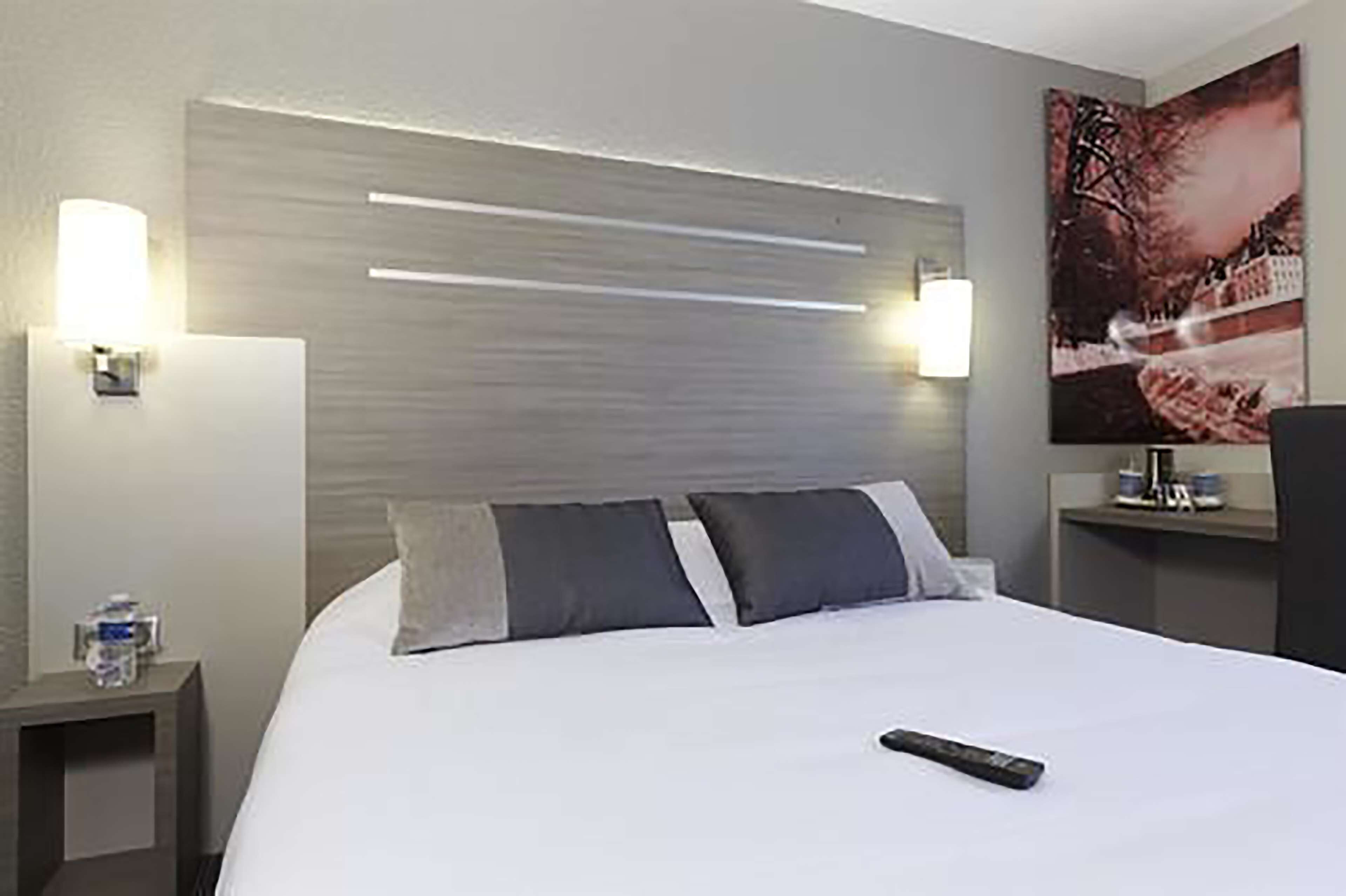 Quarto de hotel com uma cama, duas luminárias de cabeceira, uma mesinha de cabeceira, um controle remoto e um quadro na parede.