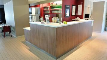 Uma área de recepção moderna com um balcão de madeira, um espaço de estar com mesa e cadeiras, e uma cozinha ao fundo.
