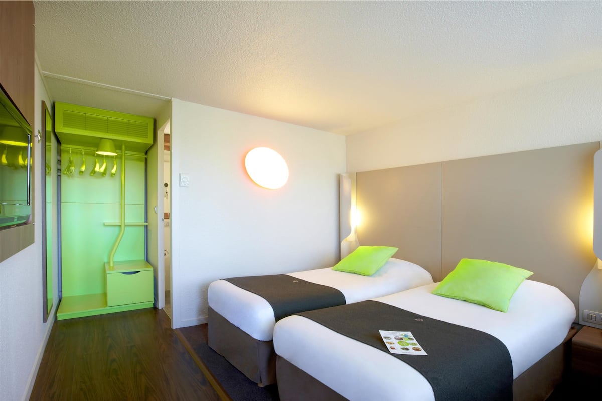 Quarto de hotel com duas camas, banheiro verde e luminária redonda na parede.