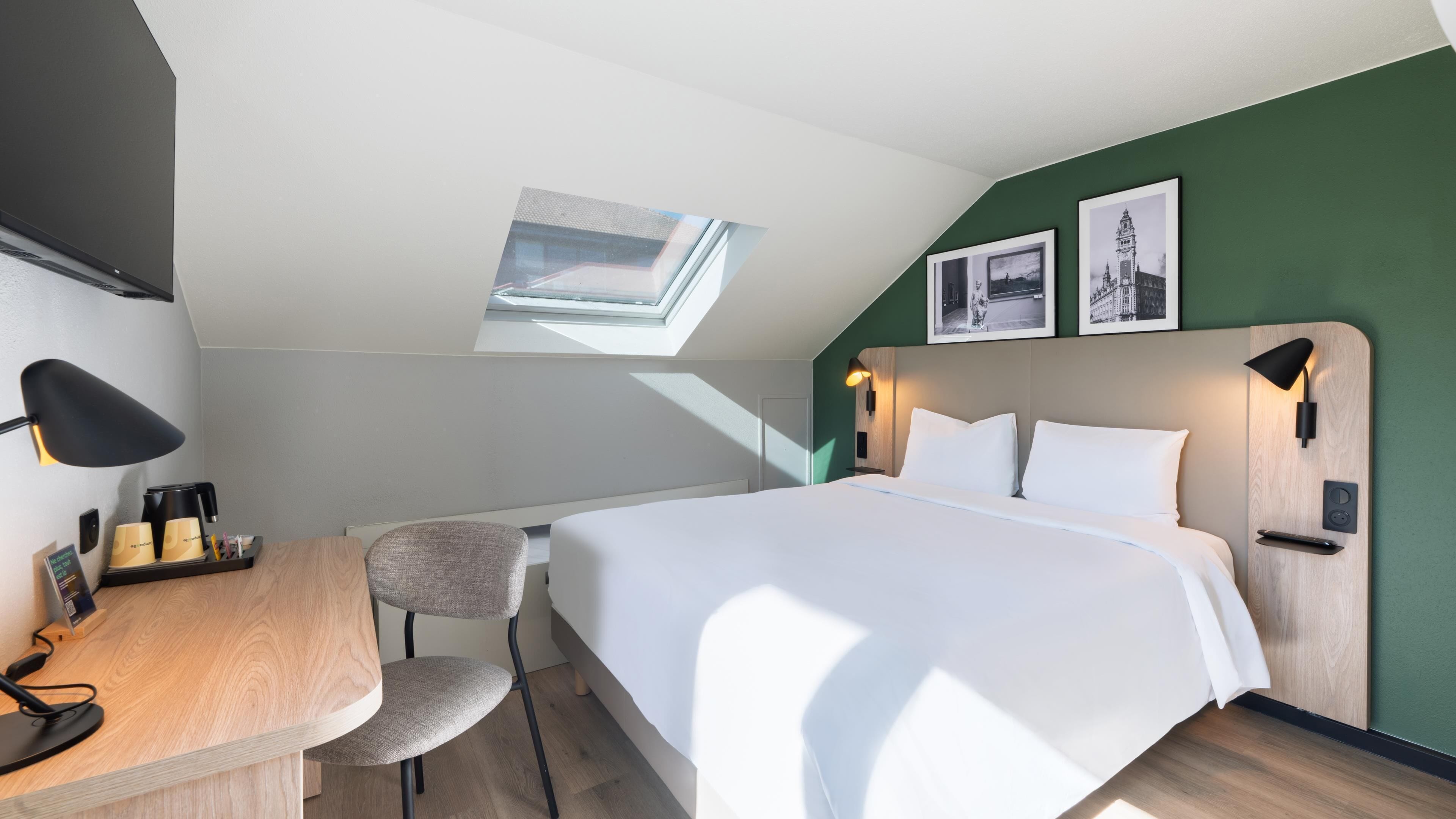 Quarto de hotel moderno com claraboia, uma cama, escrivaninha com cadeira e duas molduras na parede.