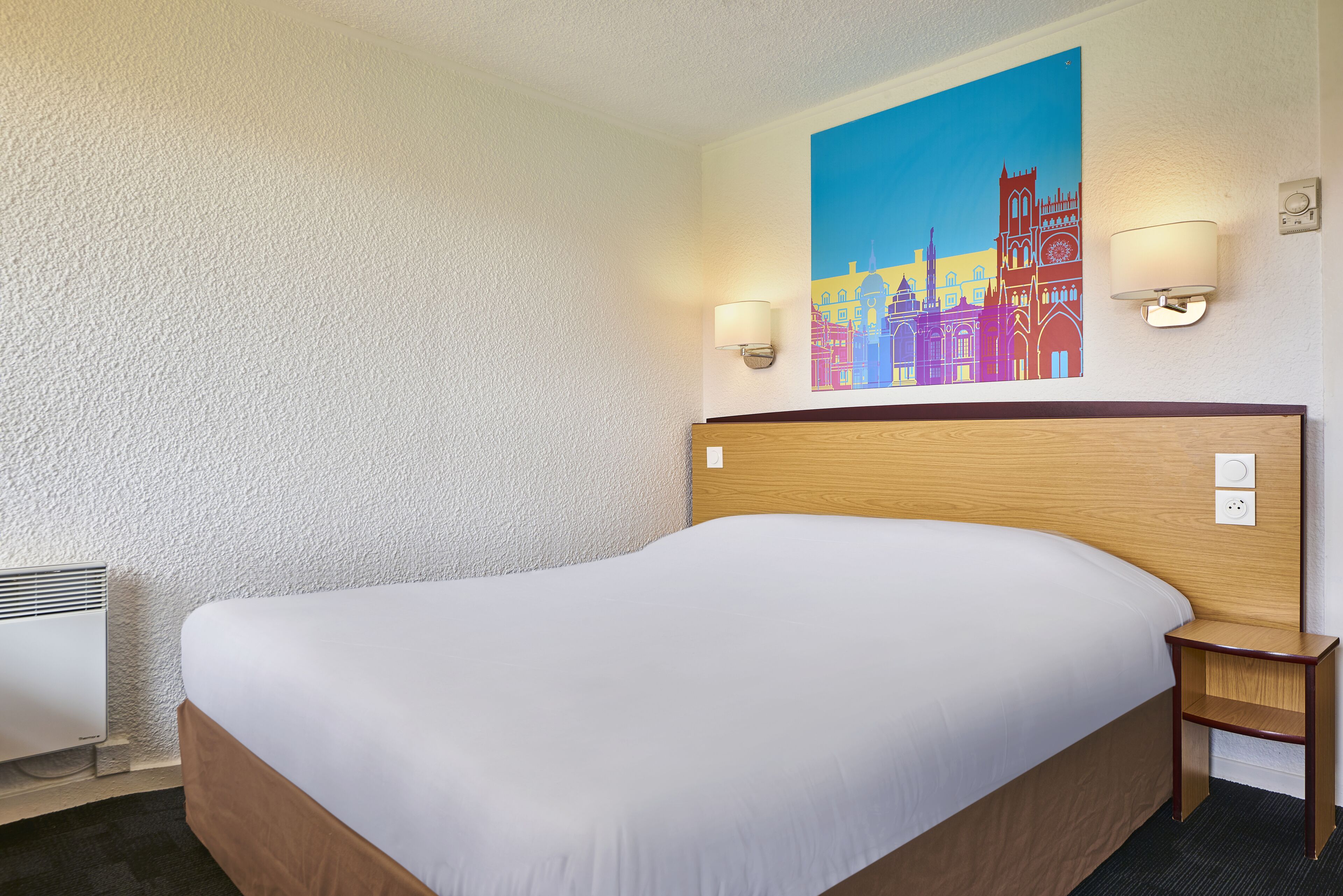 Quarto de hotel com uma cama grande, um quadro na parede, duas luminárias de cabeceira e uma pequena mesa de cabeceira de madeira.