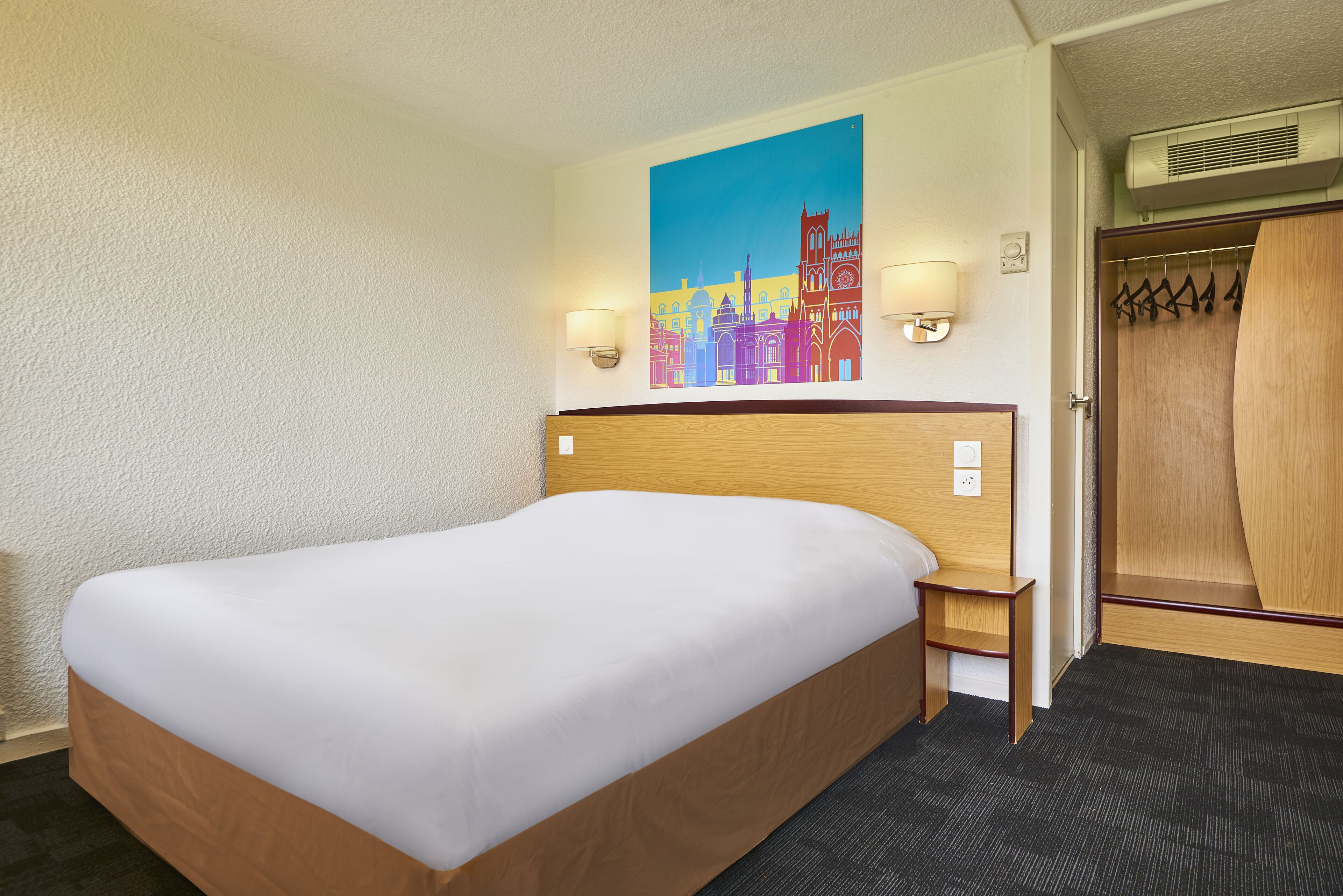 Quarto de hotel com uma cama de solteiro, cabeceira, duas luminárias fixadas na parede, pintura de paisagem urbana, ar condicionado e guarda-roupa de madeira.
