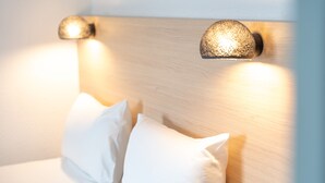 Uma cama bem arrumada com lençóis brancos e duas almofadas, uma cabeceira de madeira com uma luminária texturizada, e uma parede lisa ao fundo.