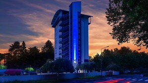 Um edifício moderno e imponente com uma característica luminosa vertical marcante, rodeado por árvores e uma estrada com rastros de luz.