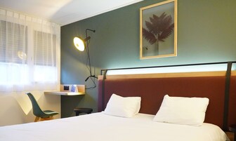 Quarto de hotel com cama, escrivaninha, cadeira, luminária na parede e planta emoldurada.