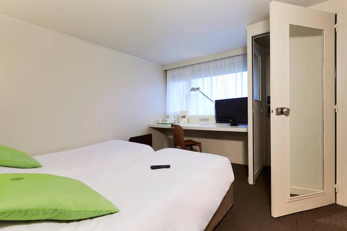 Quarto de hotel com cama, escrivaninha, cadeira, televisão e porta.