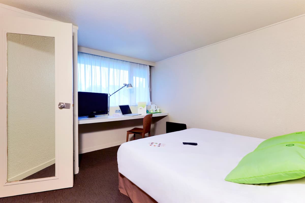 Quarto de hotel com uma cama, uma escrivaninha com computador, uma cadeira e uma janela com cortinas.