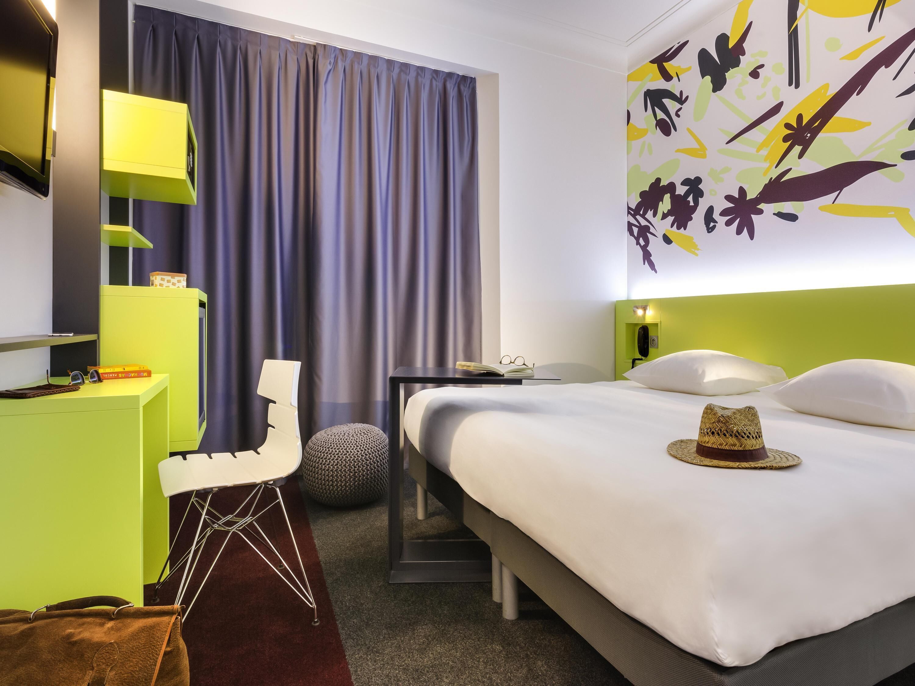 Quarto de hotel com uma cama, escrivaninha, cadeira, abajur, um banquinho redondo e uma parede com um design abstrato colorido.