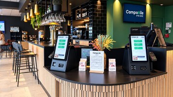 Uma área de recepção moderna com um balcão curvo, totens digitais e uma televisão com o nome do hotel.