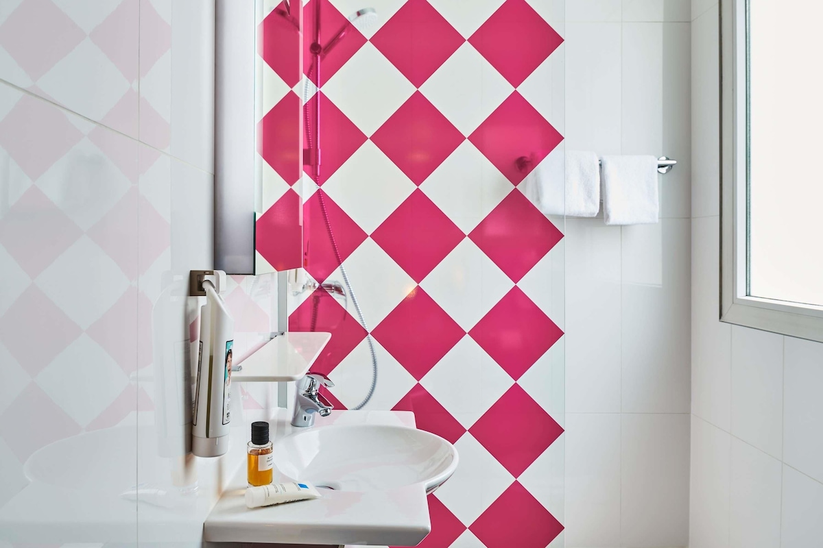 Casa de banho com lavatório branco, espelho e chuveiro com parede em padrão de losangos rosa e branco.