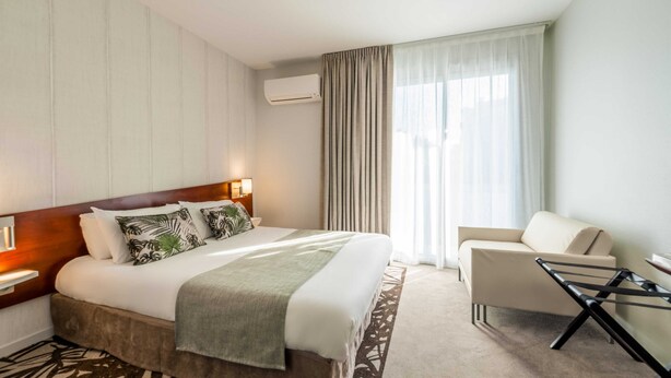 Quarto de hotel com cama, cadeira, mesa pequena, ar condicionado e janela com cortinas.