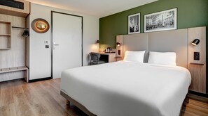 Quarto de hotel com uma cama grande, uma secretária com cadeira, um guarda-roupa e duas molduras na parede.