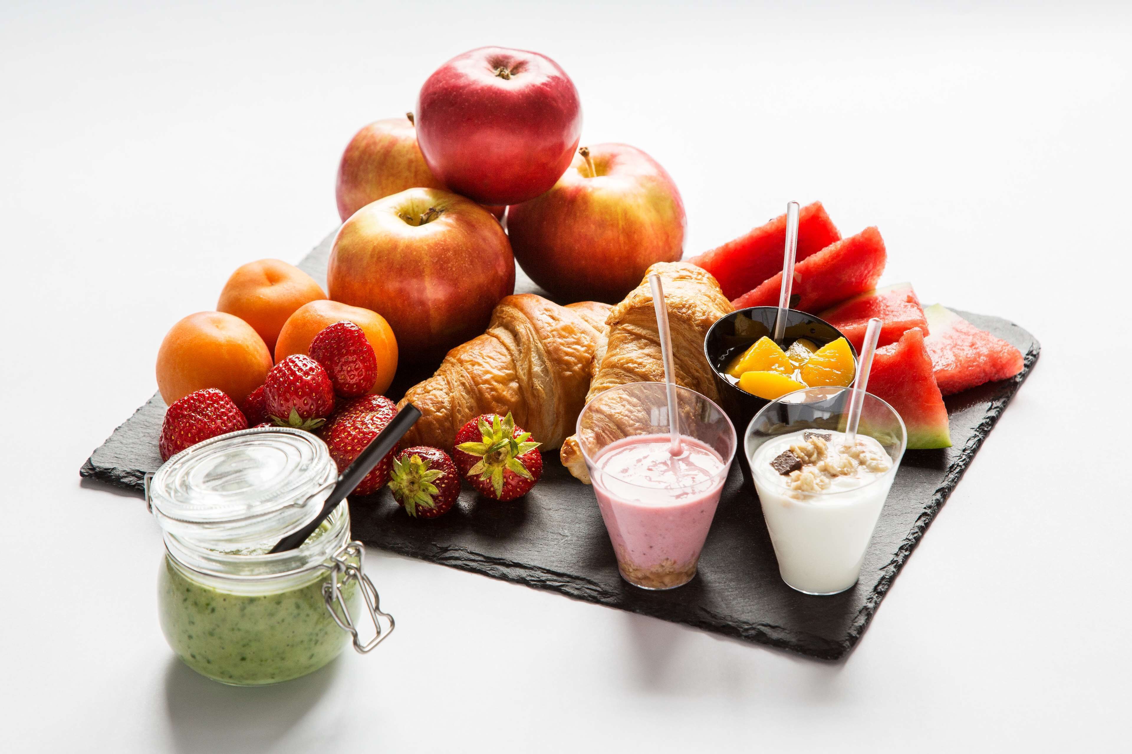 Uma mesa de café da manhã com maçãs, morangos, melancia, croissants e um smoothie.