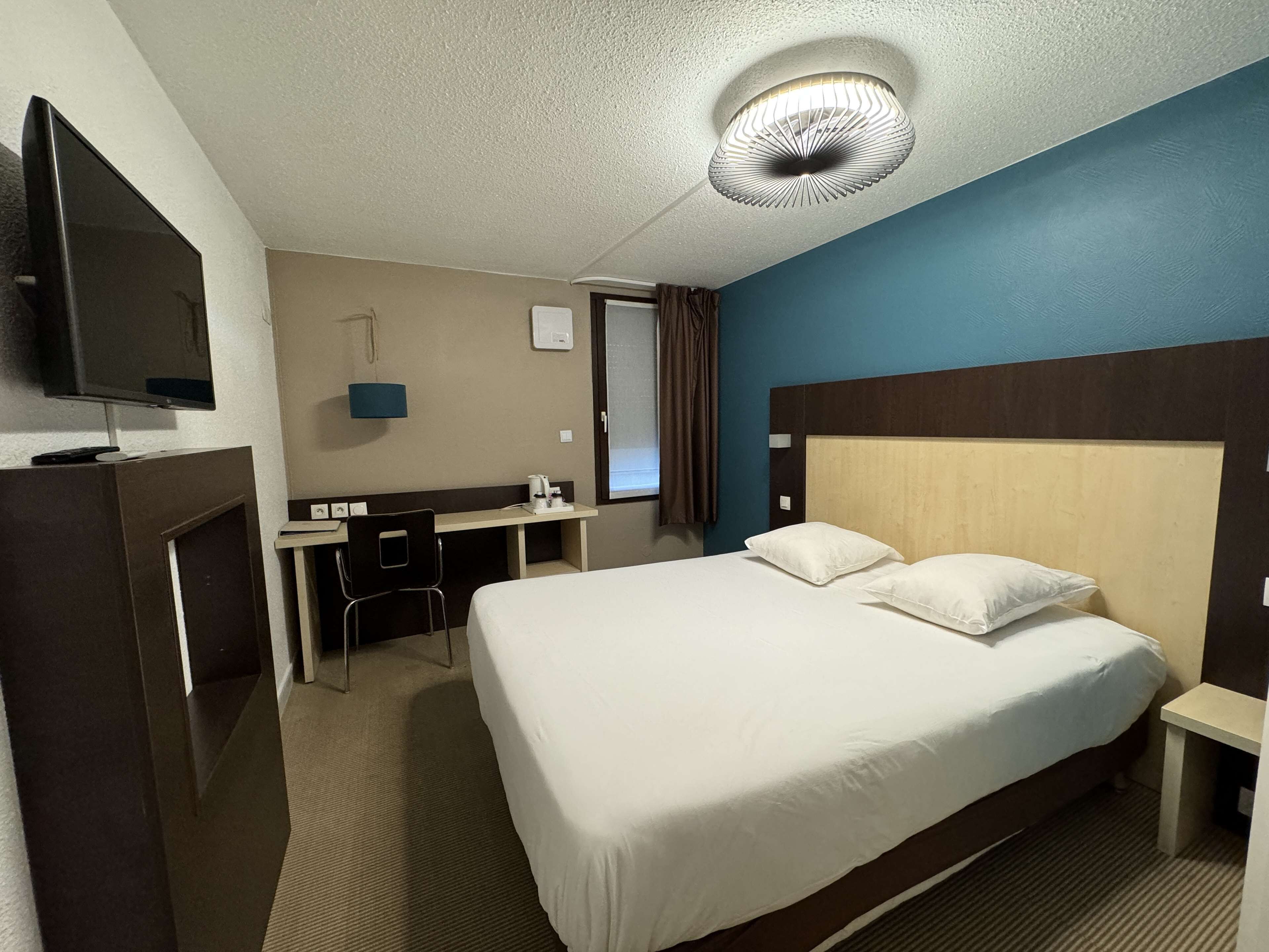 Quarto de hotel com cama, escrivaninha, cadeira, televisão e luminária no teto.