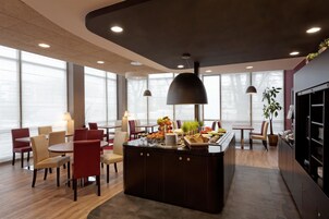 Um hall de hotel moderno com uma ilha central com buffet, assentos e grandes janelas que oferecem luz natural.