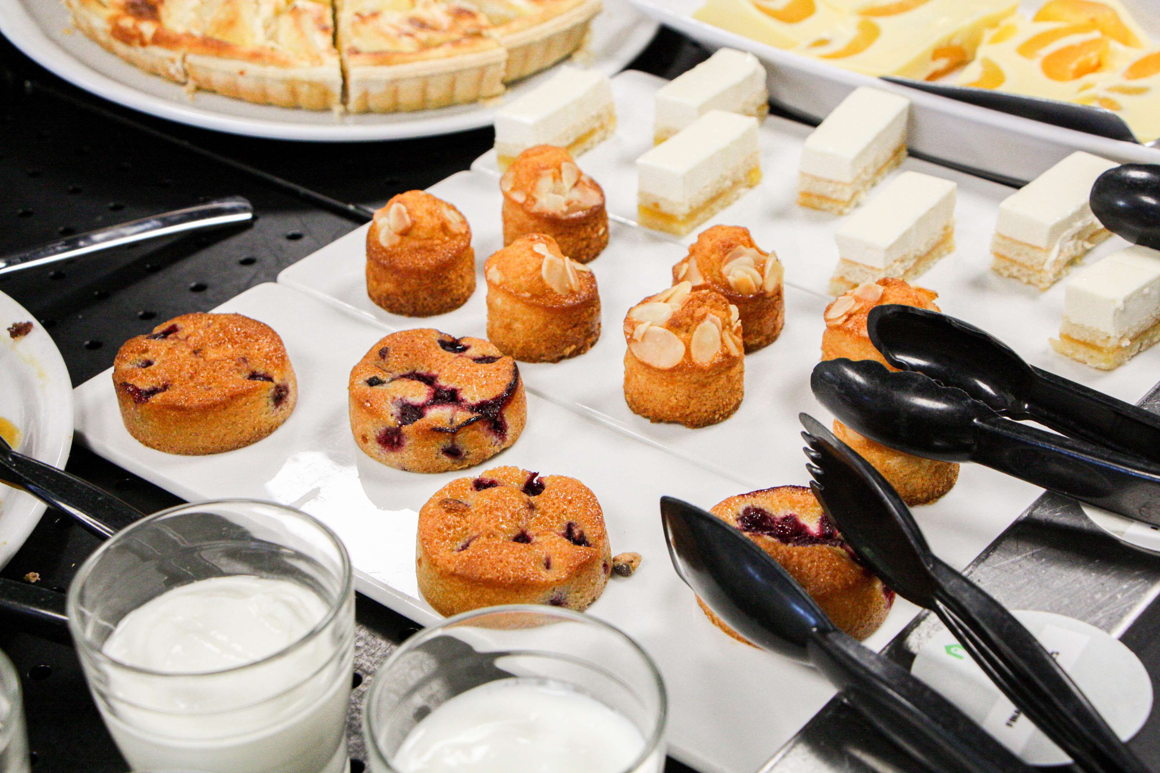 Diversos doces em um prato, incluindo muffins e bolinhos, com um conjunto de garfo e colher por perto.