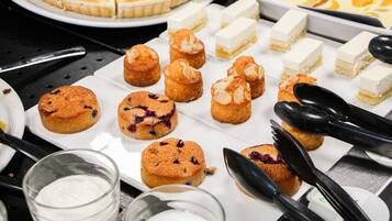 Diversos doces em um prato, incluindo muffins e bolinhos, com um conjunto de garfo e colher por perto.