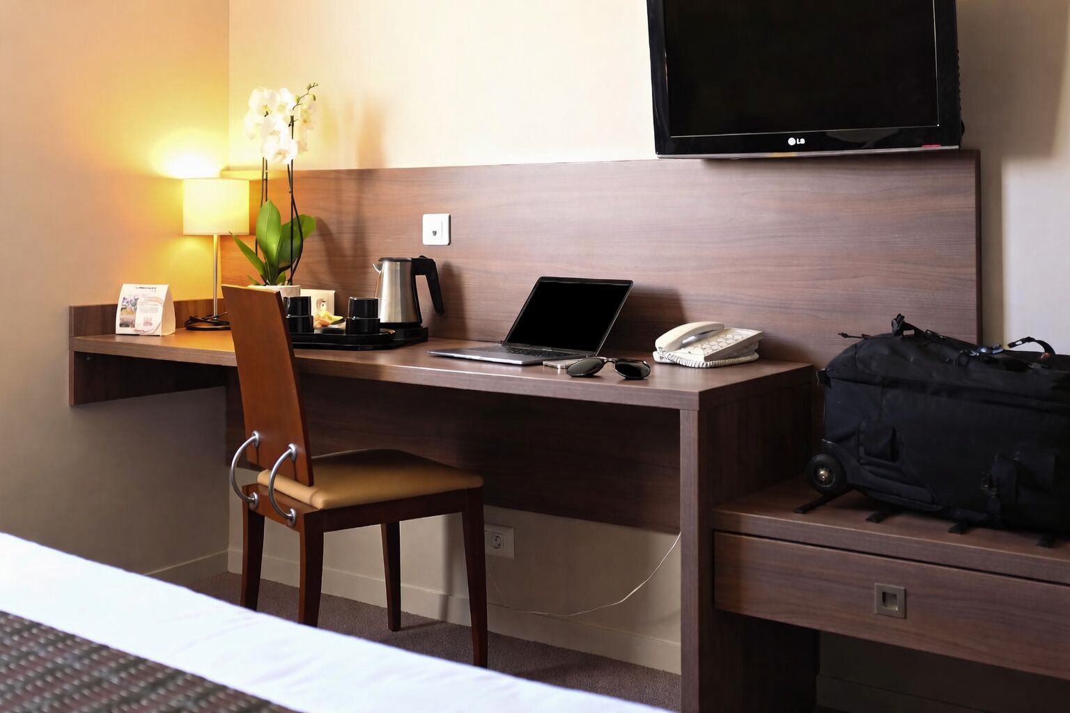 Quarto de hotel com uma escrivaninha de madeira, um laptop, um telefone, uma luminária, uma planta e uma TV fixada na parede.