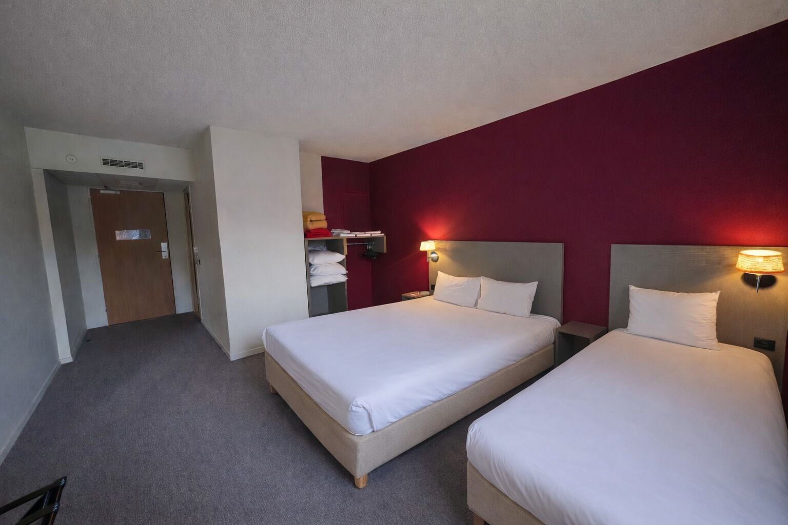 Quarto de hotel com duas camas, um guarda-roupa, uma escrivaninha e uma parede de destaque vermelha.