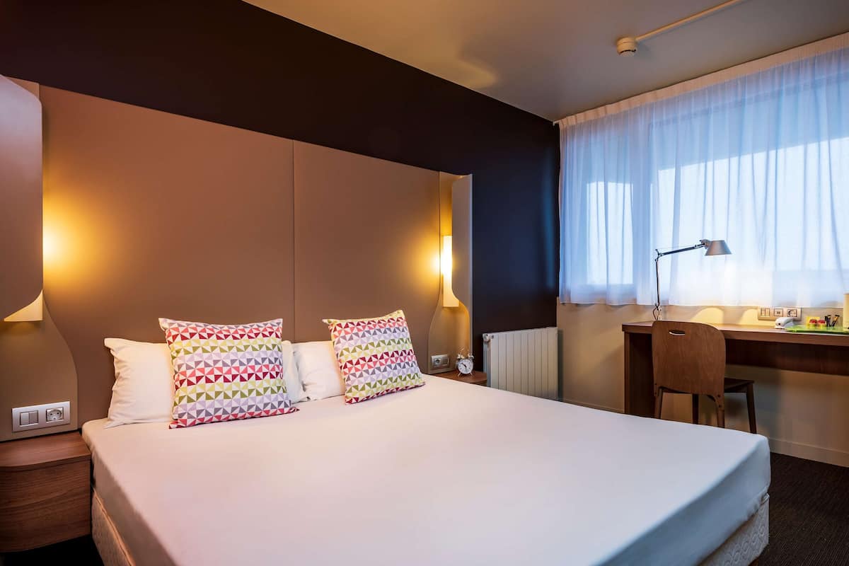 Quarto de hotel com uma cama grande, duas mesas de cabeceira com luminárias, uma escrivaninha com cadeira e uma janela com cortinas.