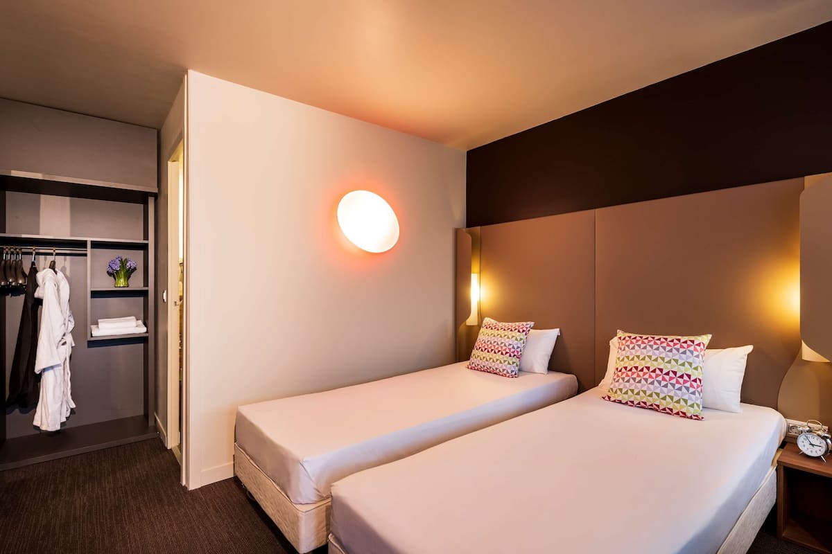 Quarto de hotel com duas camas de solteiro, um closet espaçoso e uma luminária redonda na parede.