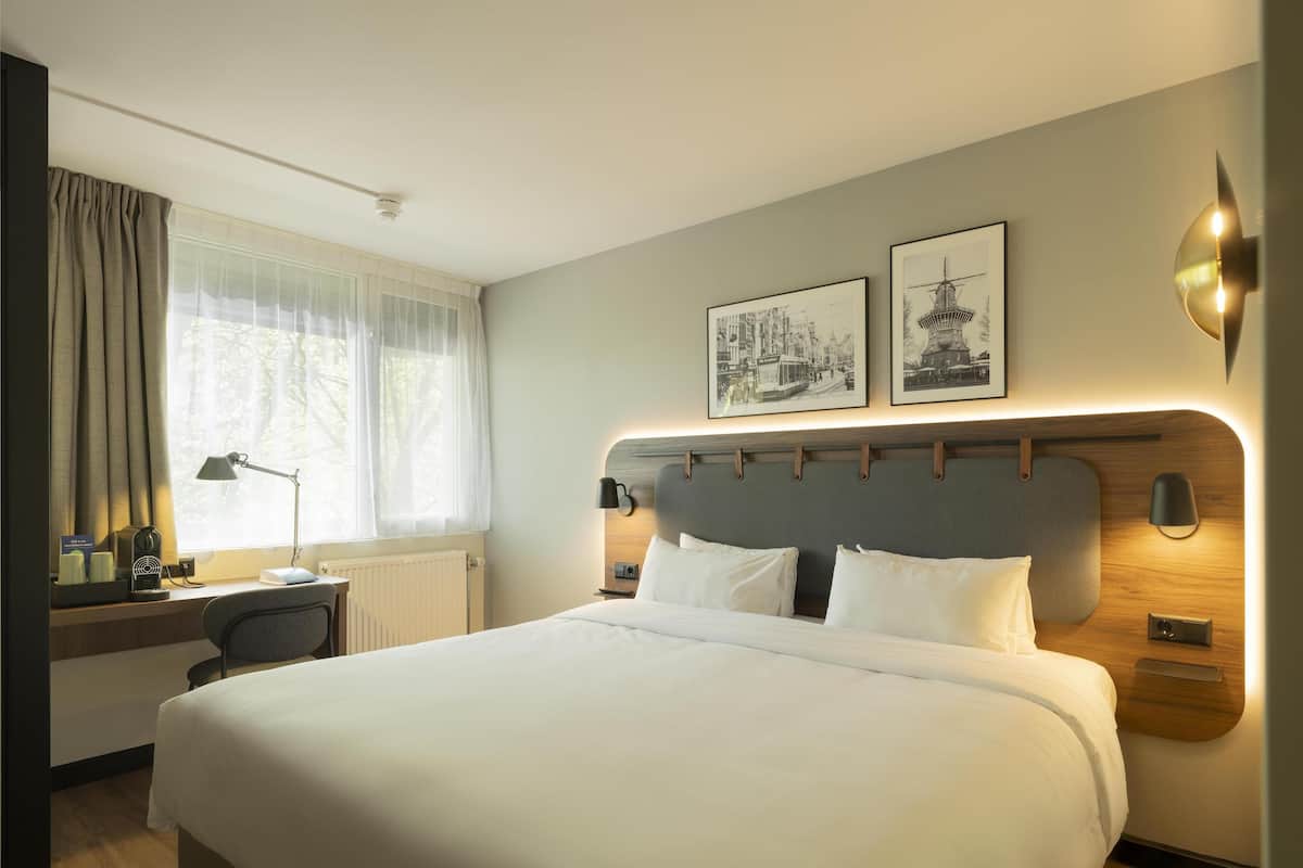 Quarto de hotel com uma cama grande, uma escrivaninha com abajur, uma janela com cortinas e duas molduras na parede.