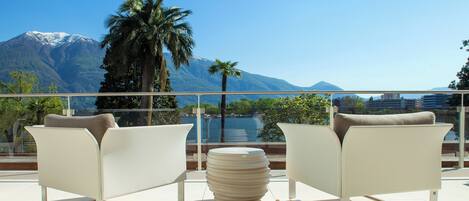 Terraço com cadeiras de descanso brancas e uma mesa pequena, com vista para uma piscina e montanhas.