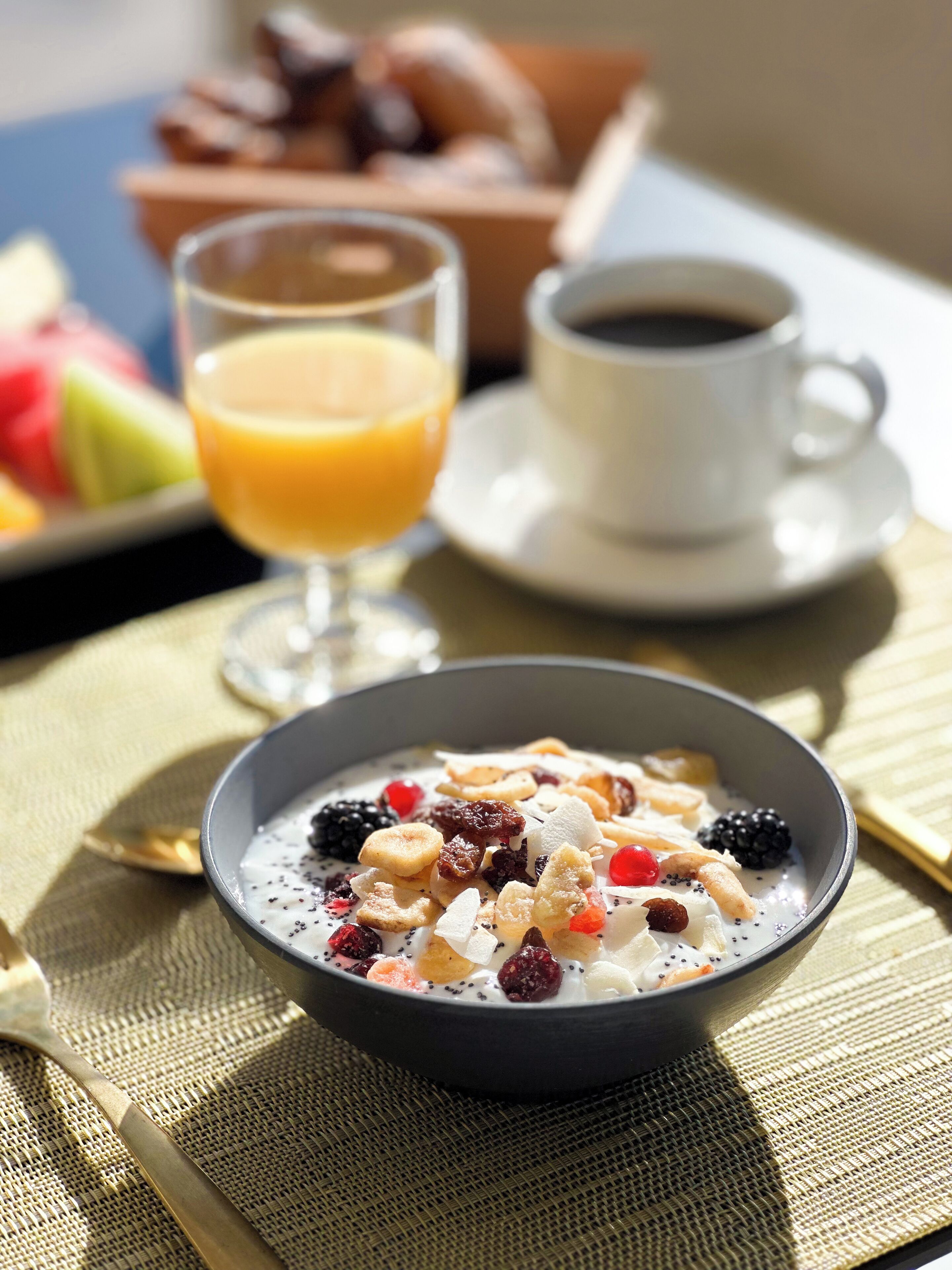 Um ambiente de café da manhã com uma tigela de iogurte coberta com frutas e nozes, um copo de suco de laranja e uma xícara de café sobre uma mesa.