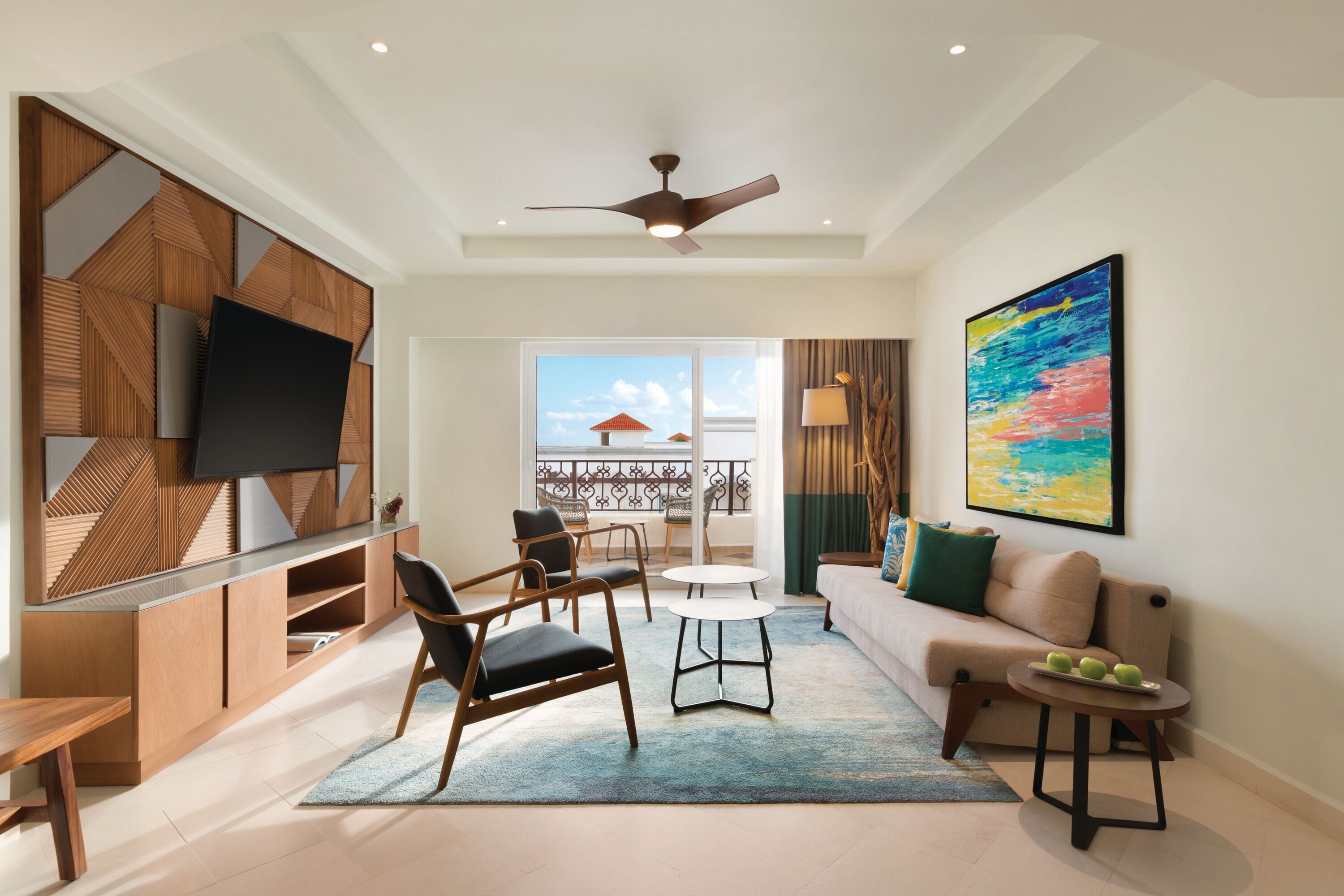 Uma sala de estar moderna com um sofá, cadeiras, uma mesa de centro e uma televisão. Há uma varanda com guarda-corpo e vista para o céu.