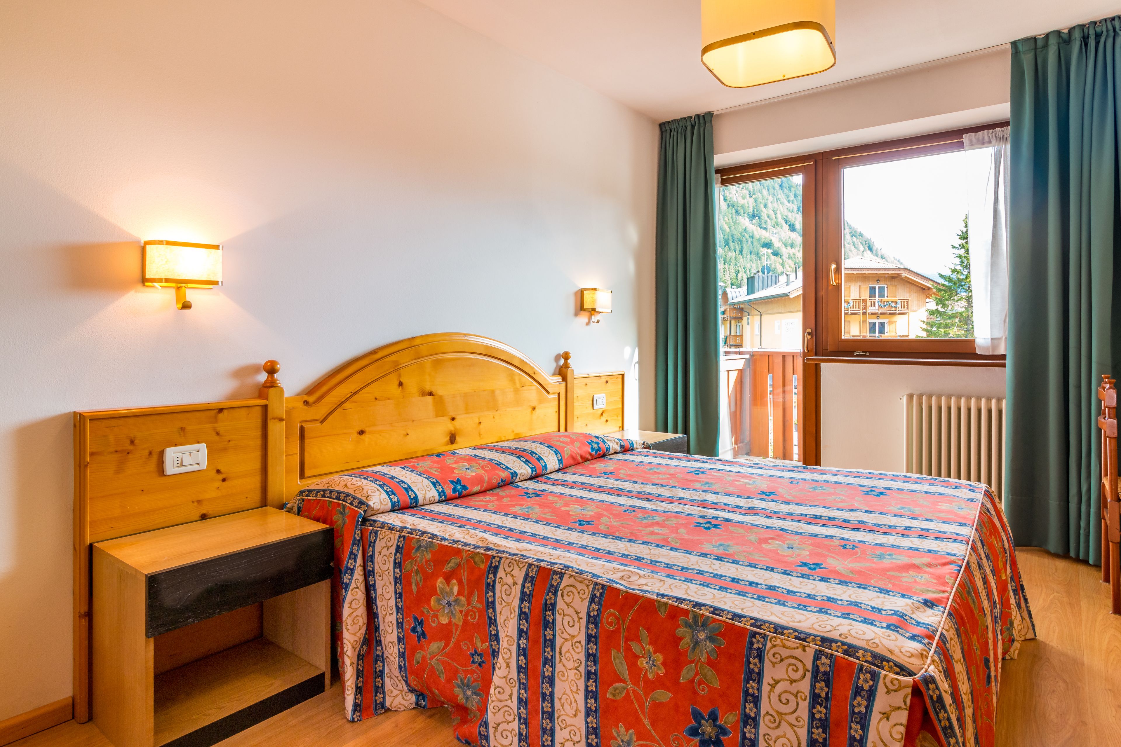 Quarto com cabeceira de madeira, cama com colcha estampada, abajures e janela com cortinas.