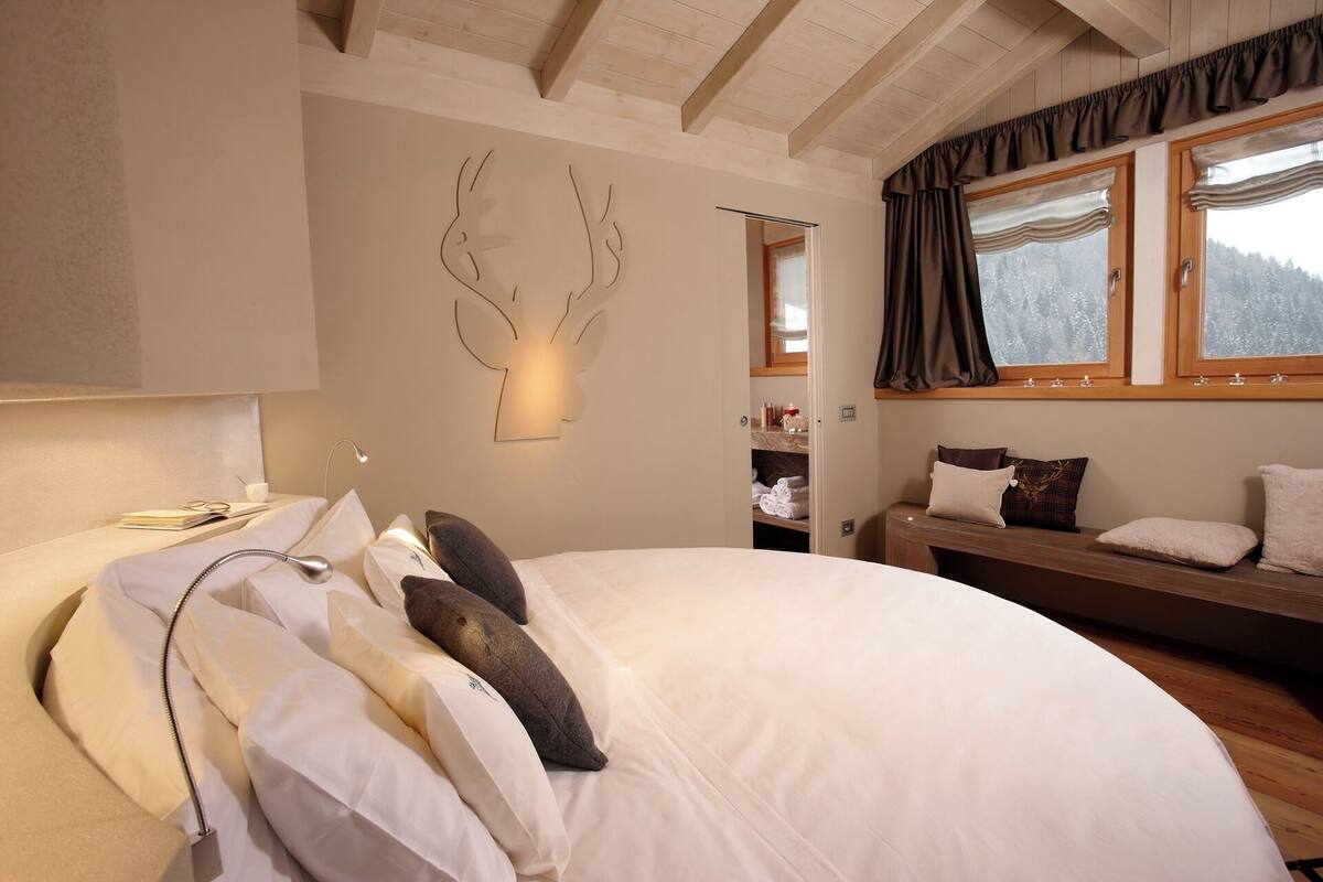 Quarto com cama, mesa de cabeceira, abajur, arte na parede, janela com cortinas e vista para árvores.